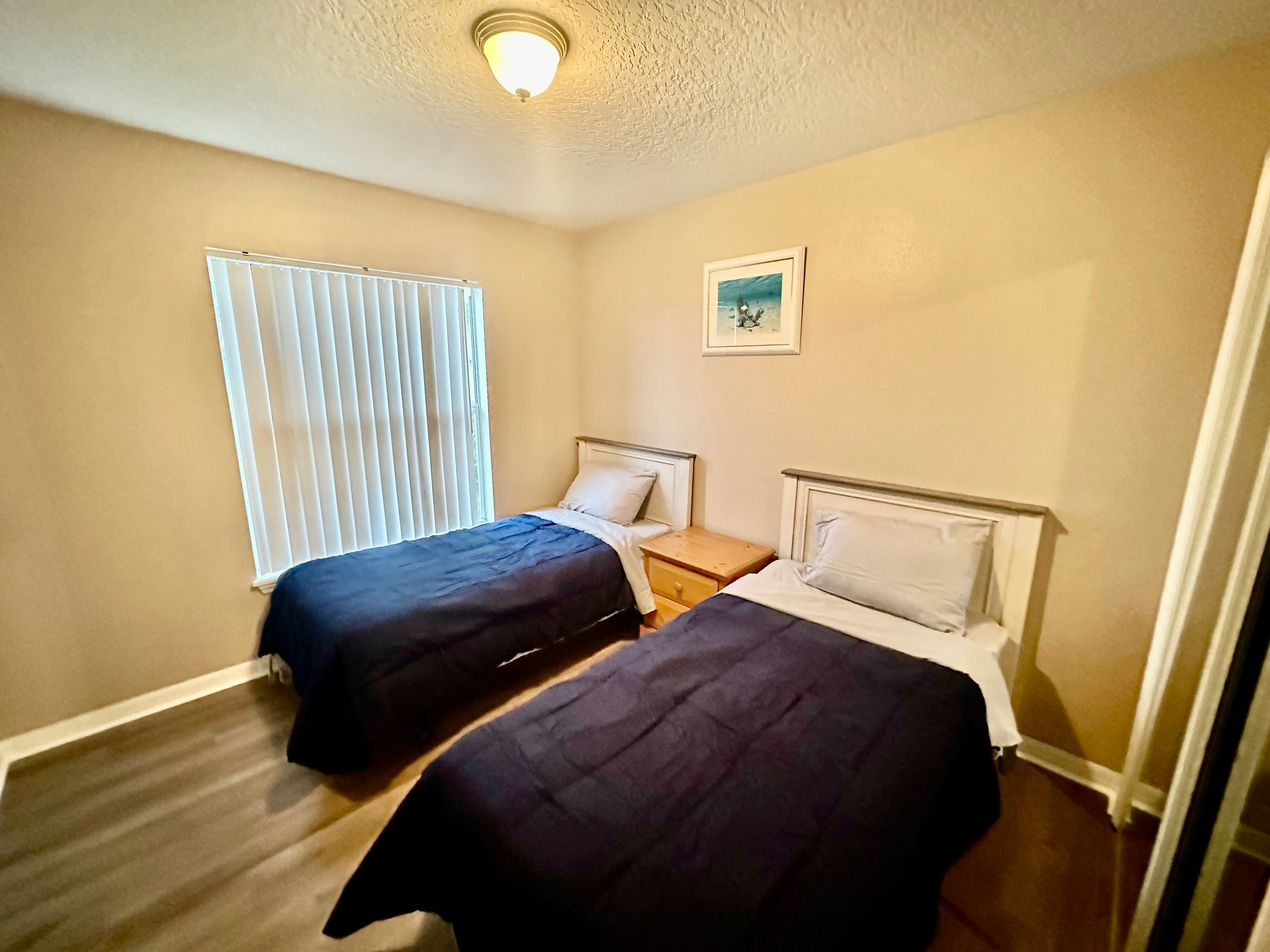 Quarto com duas camas, janela com persianas, mesa de cabeceira e luminária no teto.