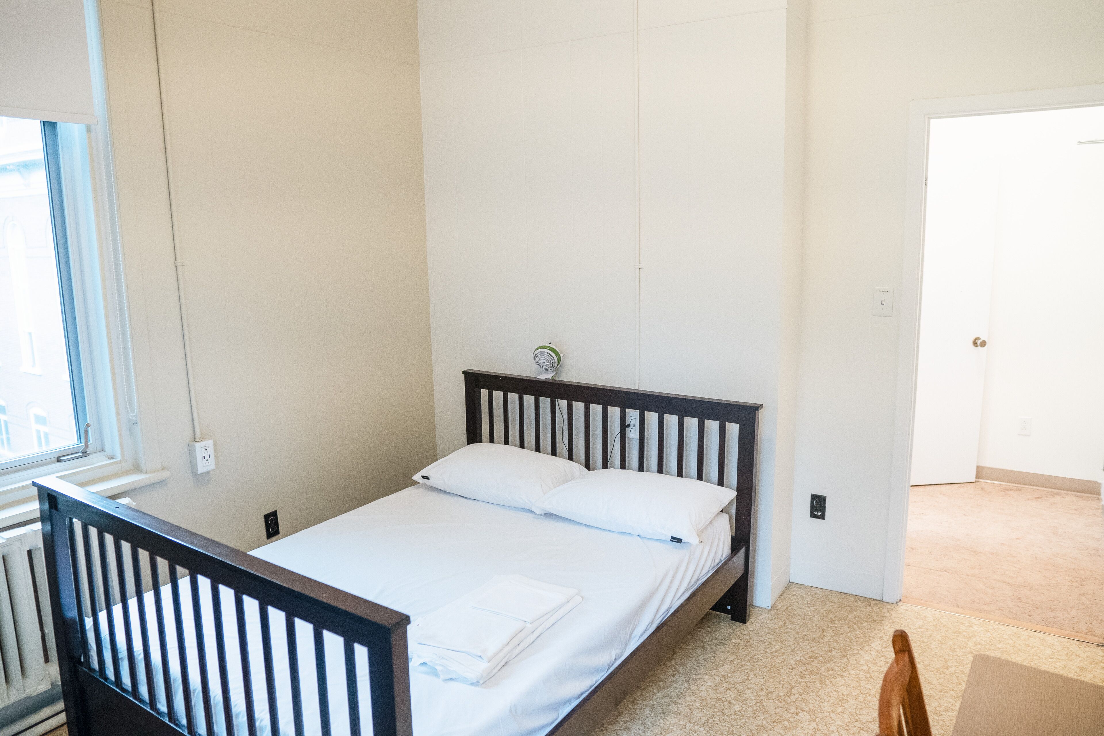 Uma cama de solteiro com cabeceira de madeira em quarto com paredes lisas e chão coberto por carpete.