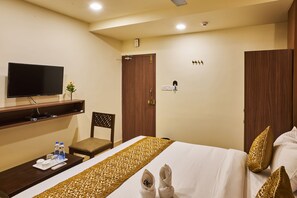 Quarto de hotel com uma cama, televisão em uma prateleira, uma cadeira, escrivaninha com artigos de higiene e um guarda-roupa.
