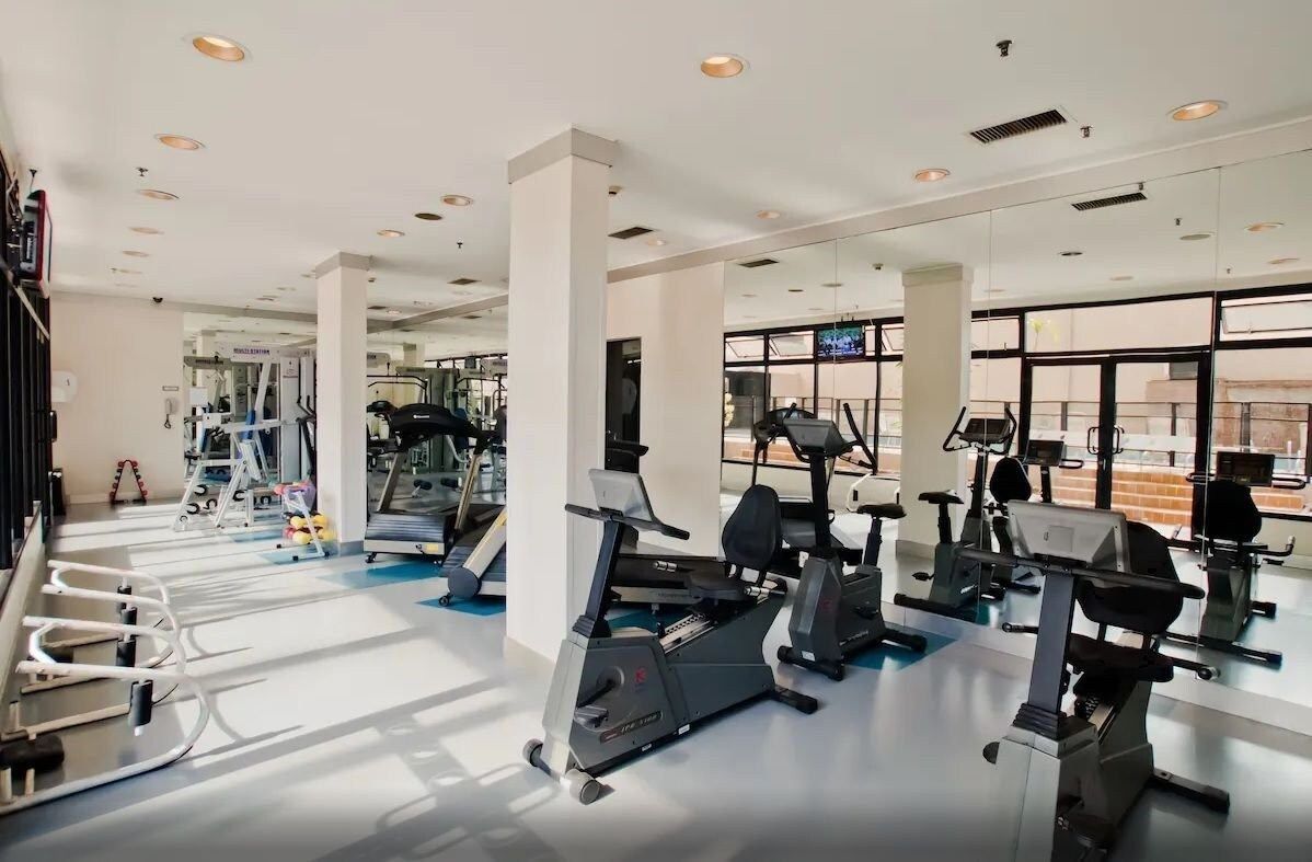Uma academia com diversos equipamentos de exercício, incluindo esteiras e elípticos, dispostos em um espaço amplo com janelas grandes e iluminação natural.