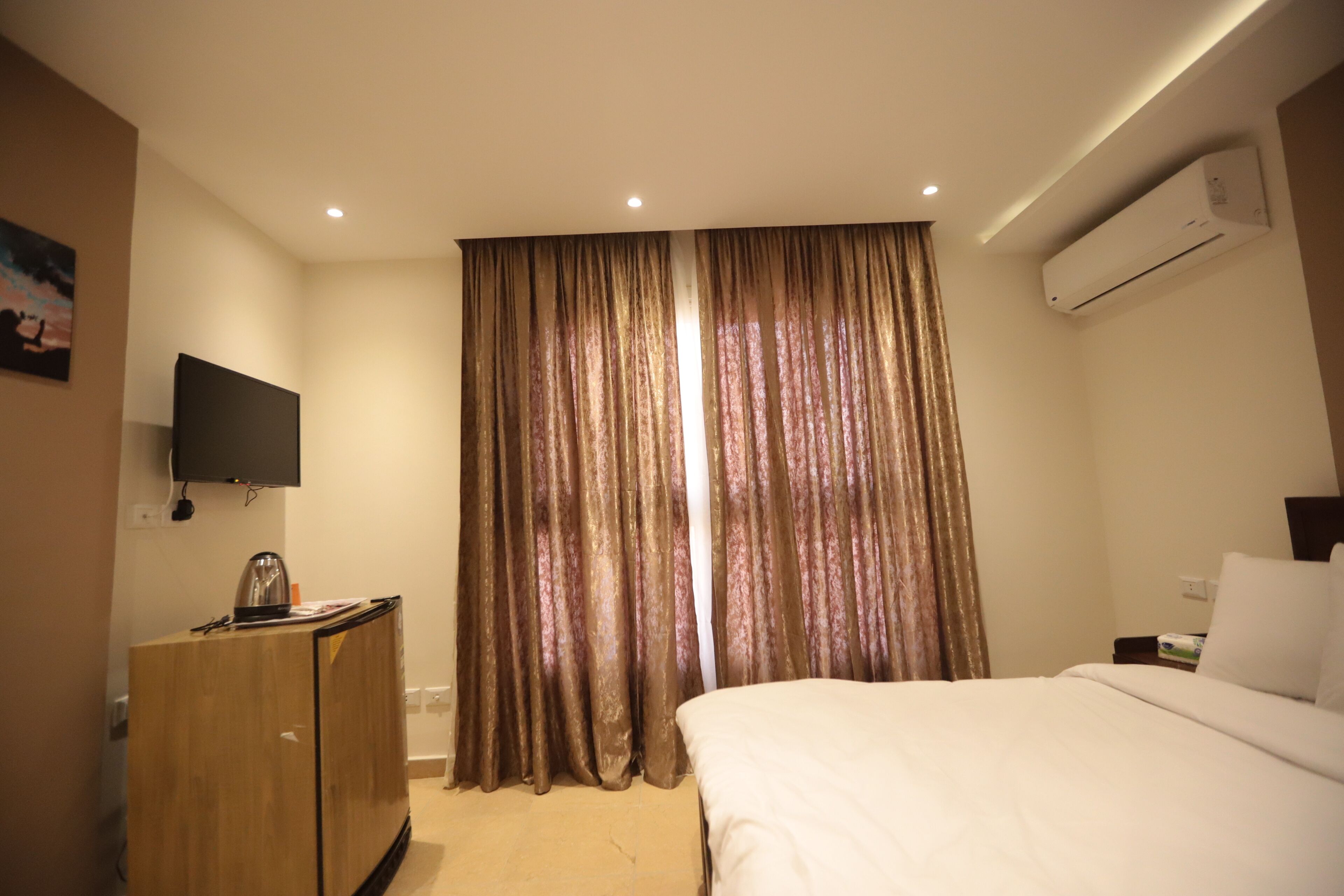 Quarto de hotel com cama, televisão na parede, cómoda de madeira e janela com cortinas.