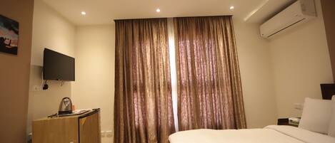 Quarto de hotel com cama, televisão na parede, cómoda de madeira e janela com cortinas.