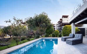 Piscina com deck revestido, rodeada por vegetação e um edifício moderno com varandas.