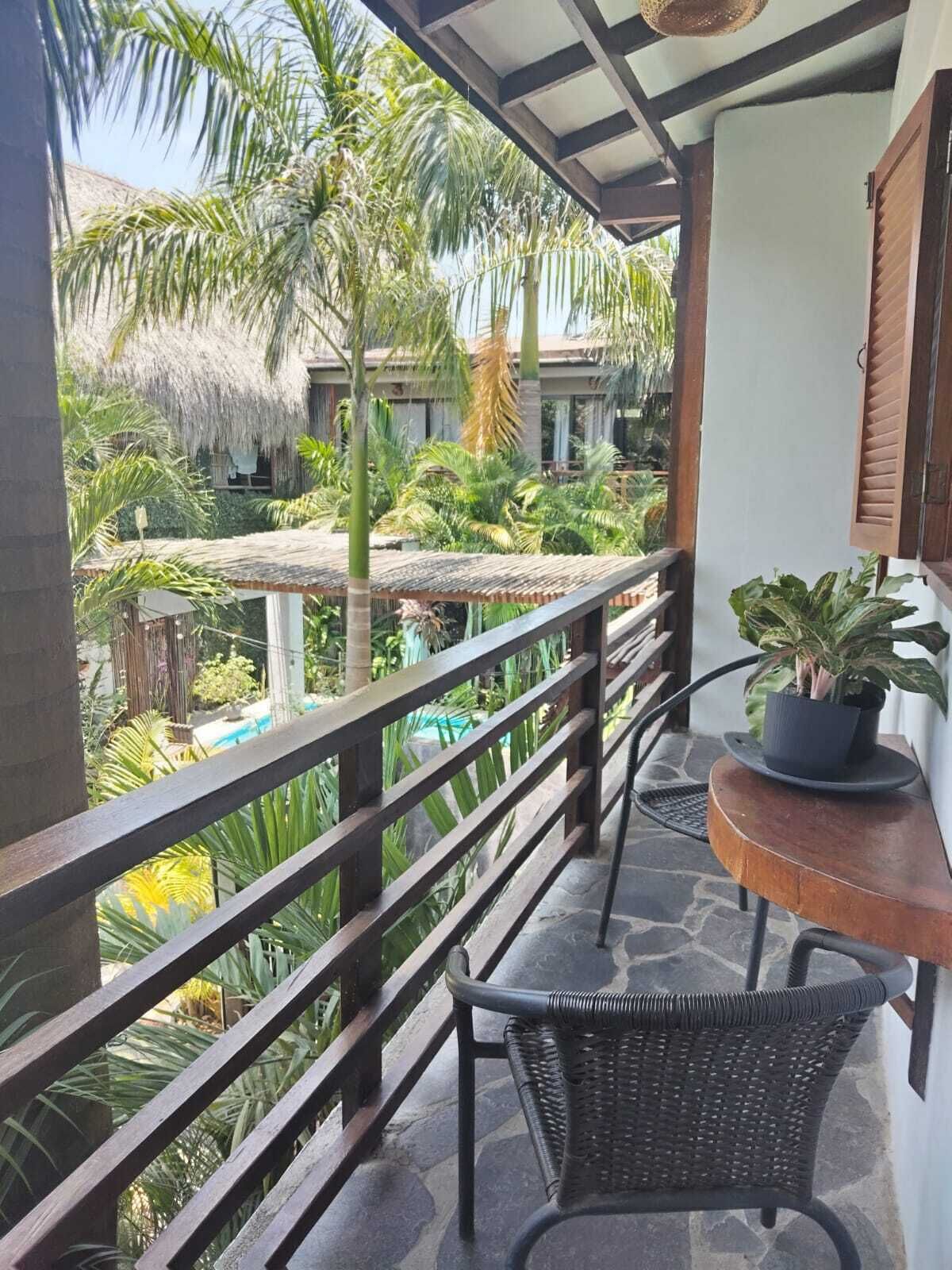 Uma varanda com vista para uma piscina e plantas tropicais, uma cadeira de vime e uma mesa de madeira com um vaso de planta.