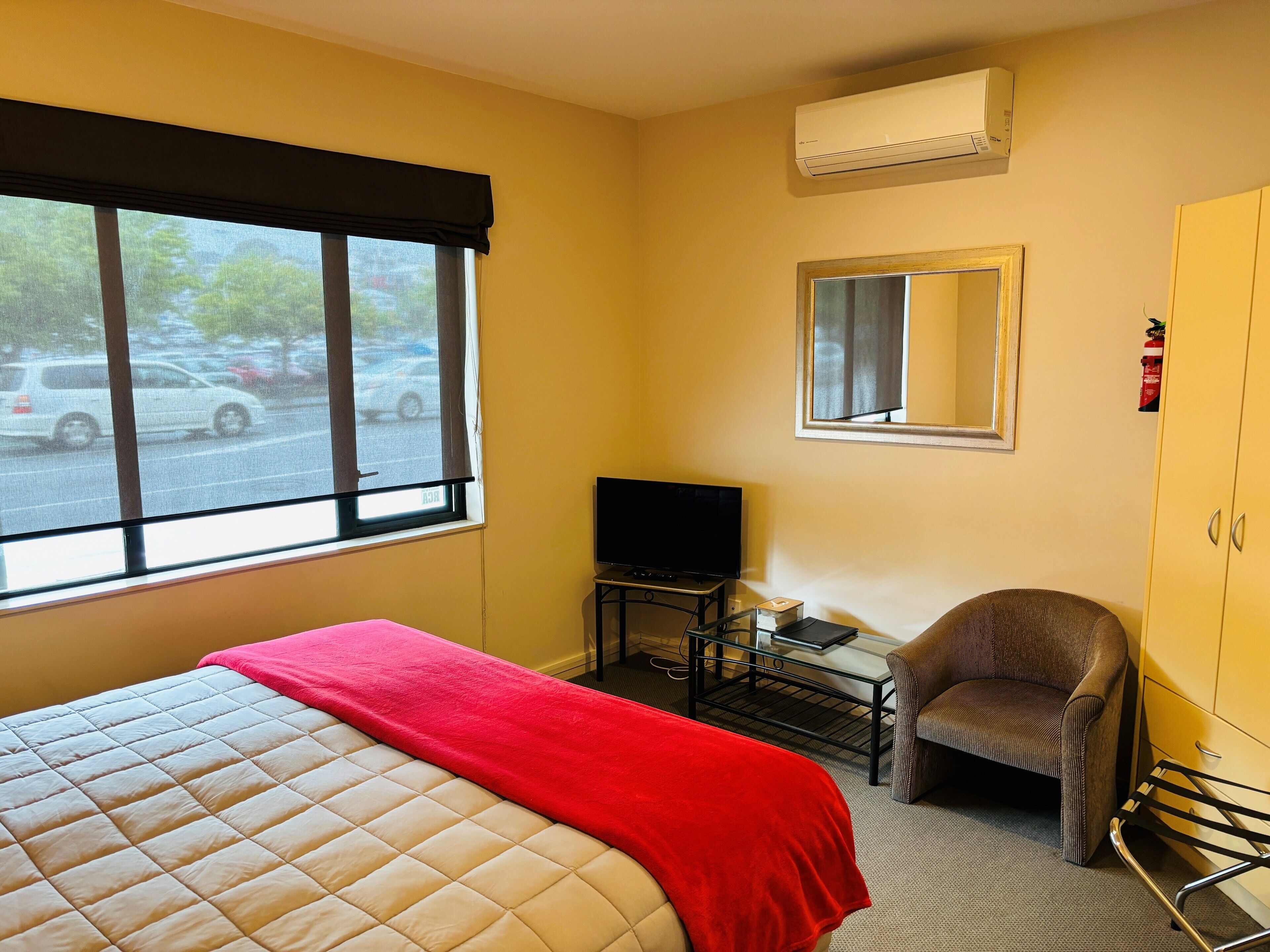 Quarto de hotel com cama, cadeira, televisão e janela com vista para carros estacionados.