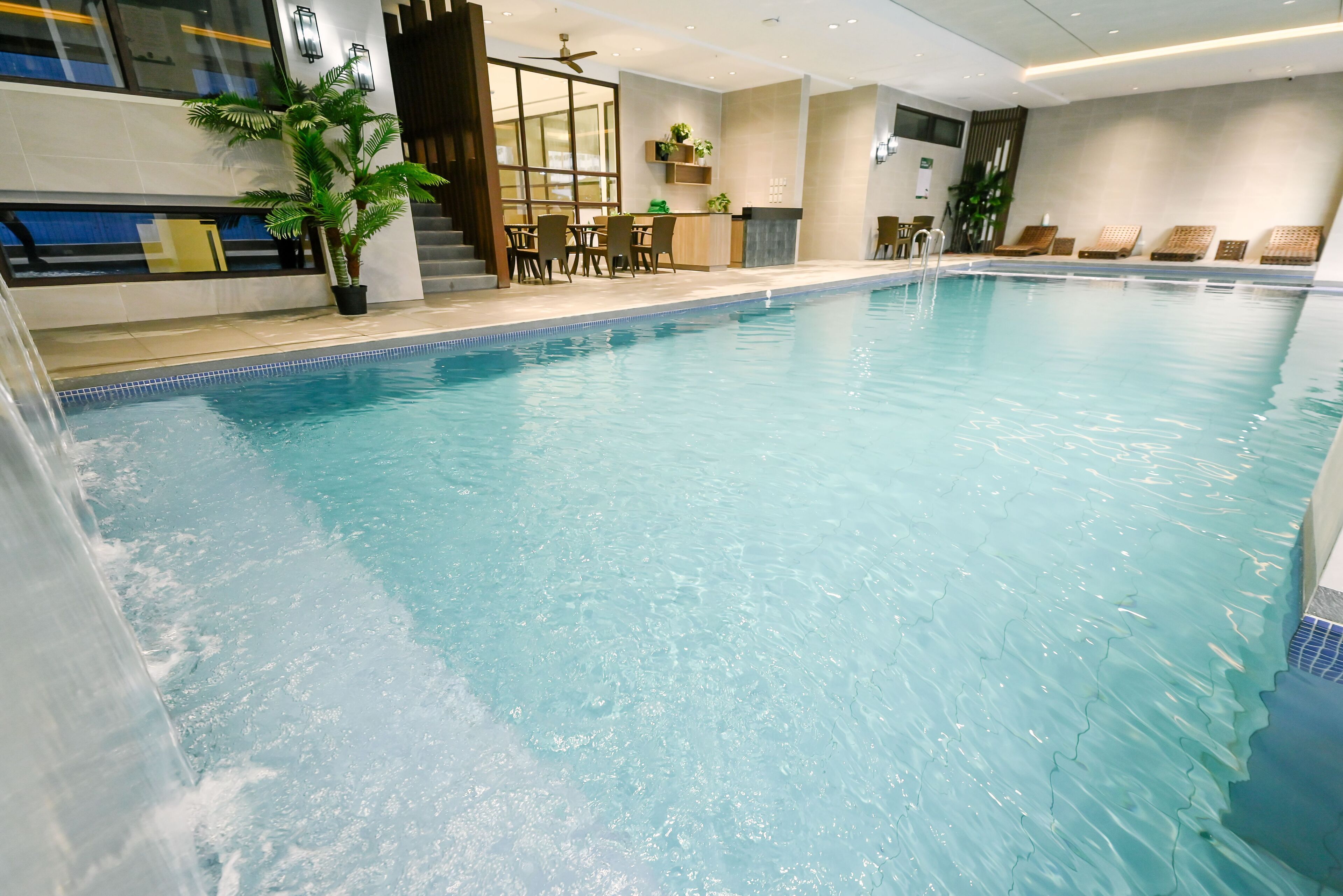 Piscina interior com água cristalina, rodeada por um interior moderno com área de estar, escada e bancada de cozinha.