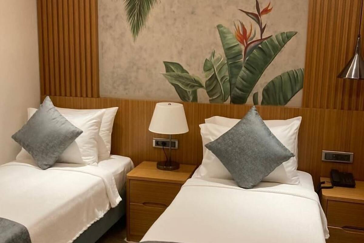 Quarto de hotel com duas camas, uma mesa de cabeceira com abajur, um mural na parede e uma cabeceira de madeira.