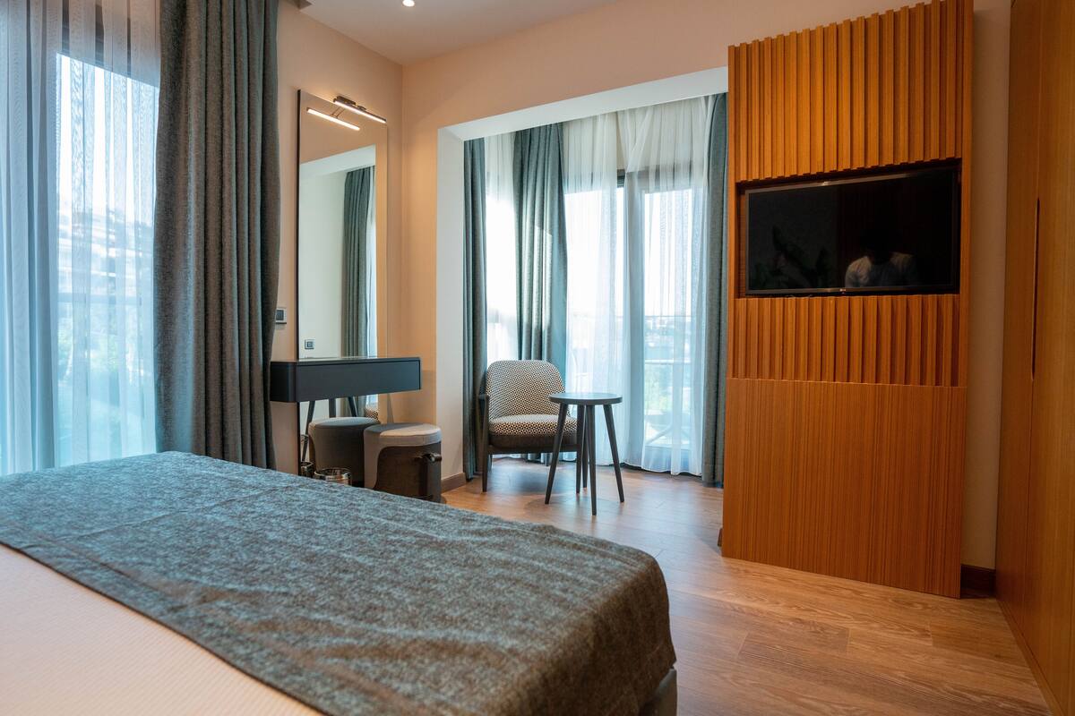 Quarto de hotel com cama grande, escrivaninha, cadeira, televisão e janela com cortinas.
