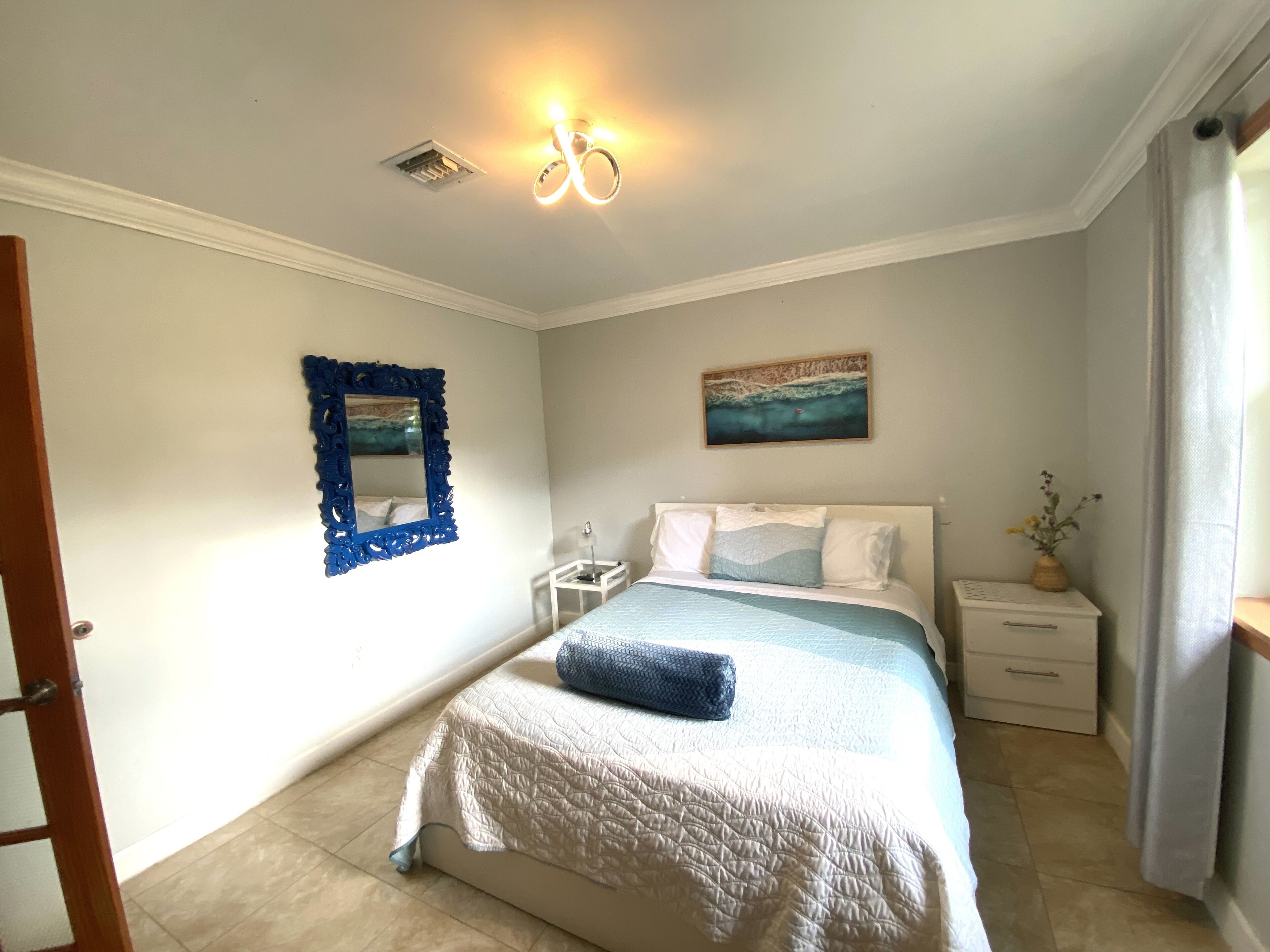Quarto com cama, mesas de cabeceira, espelho, quadro e janela com cortinas.