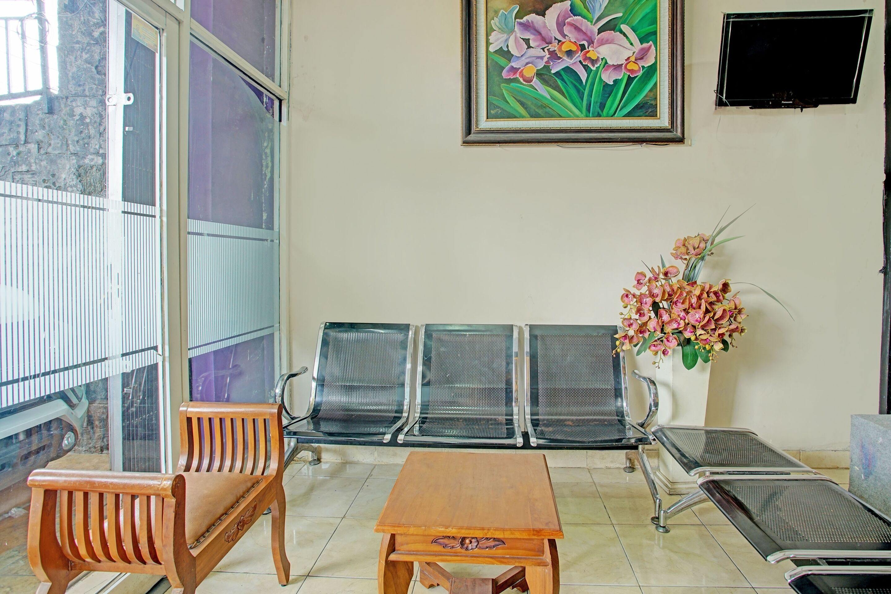 Uma área de espera com um banco de madeira, uma mesa pequena e uma cadeira. Há três cadeiras de metal e um vaso com flores. Um quadro de flores está na parede, e uma TV está instalada acima das cadeiras.