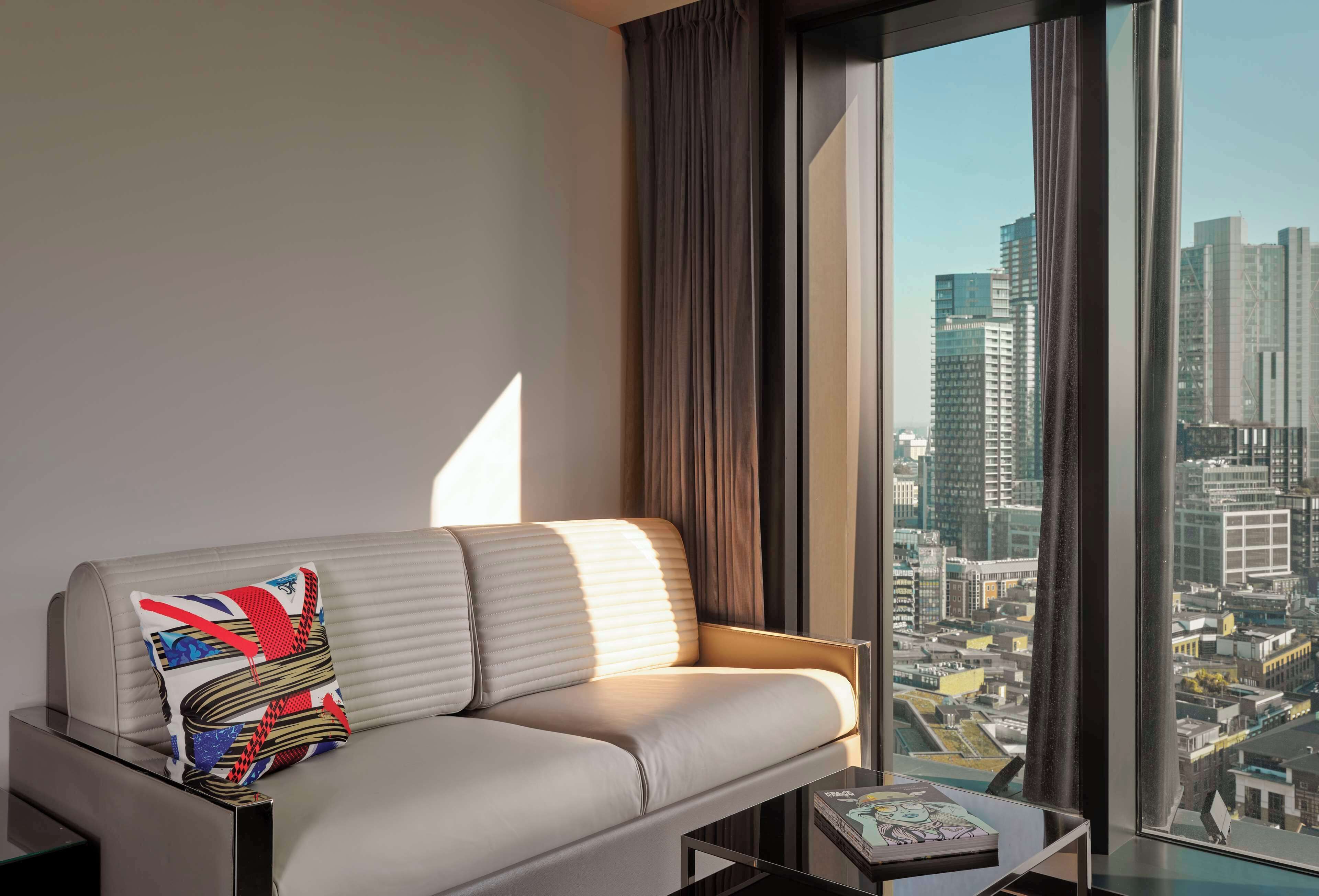 Quarto de hotel moderno com sofá e mesa de centro, janelas amplas com vista para a cidade.