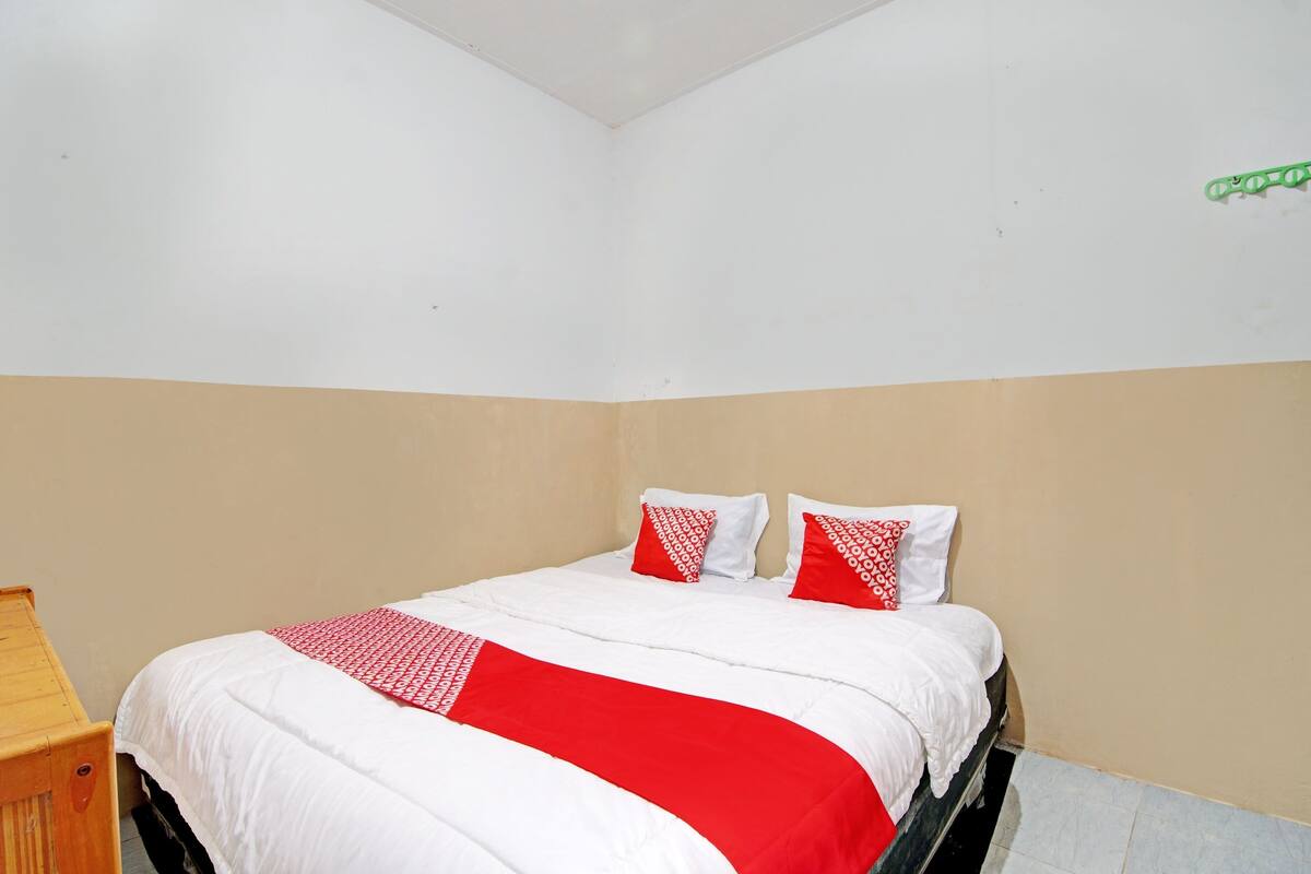 Cama bem arrumada com colcha vermelha e branca, duas almofadas com padrão vermelho e branco e uma mesa de cabeceira de madeira à esquerda.