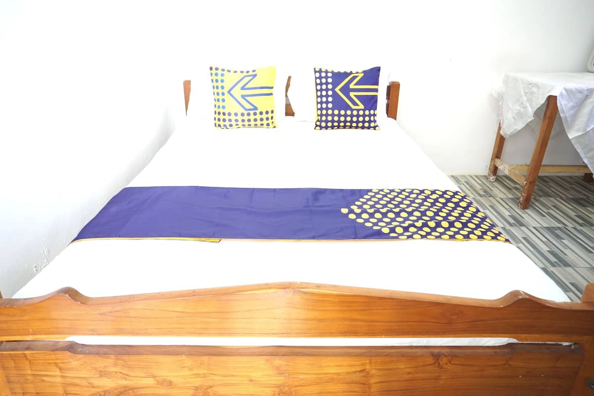 Uma cama com estrutura de madeira, roupa de cama branca e duas almofadas com padrões geométricos.