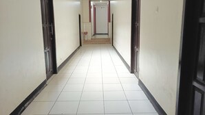 Um corredor comprido e estreito com pisos e paredes de azulejos brancos, com portas escuras de cada lado.
