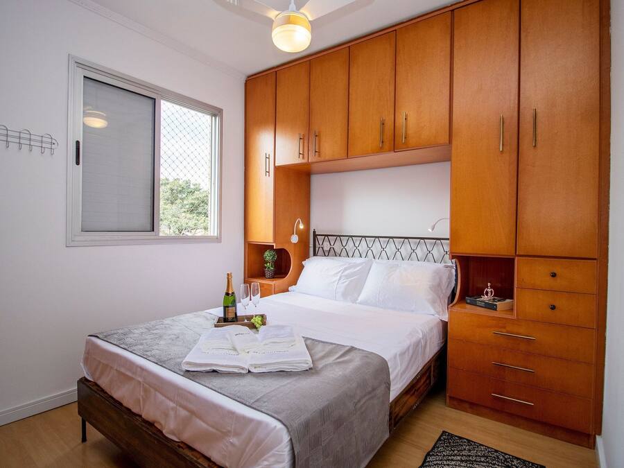 Quarto com cama, guarda-roupa de madeira e uma mesinha com garrafa e copos.