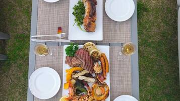 Uma montagem para piquenique com uma mesa posta para quatro pessoas, exibindo diversos tipos de carne grelhada, frutos do mar e legumes, acompanhados de copos de vinho branco.