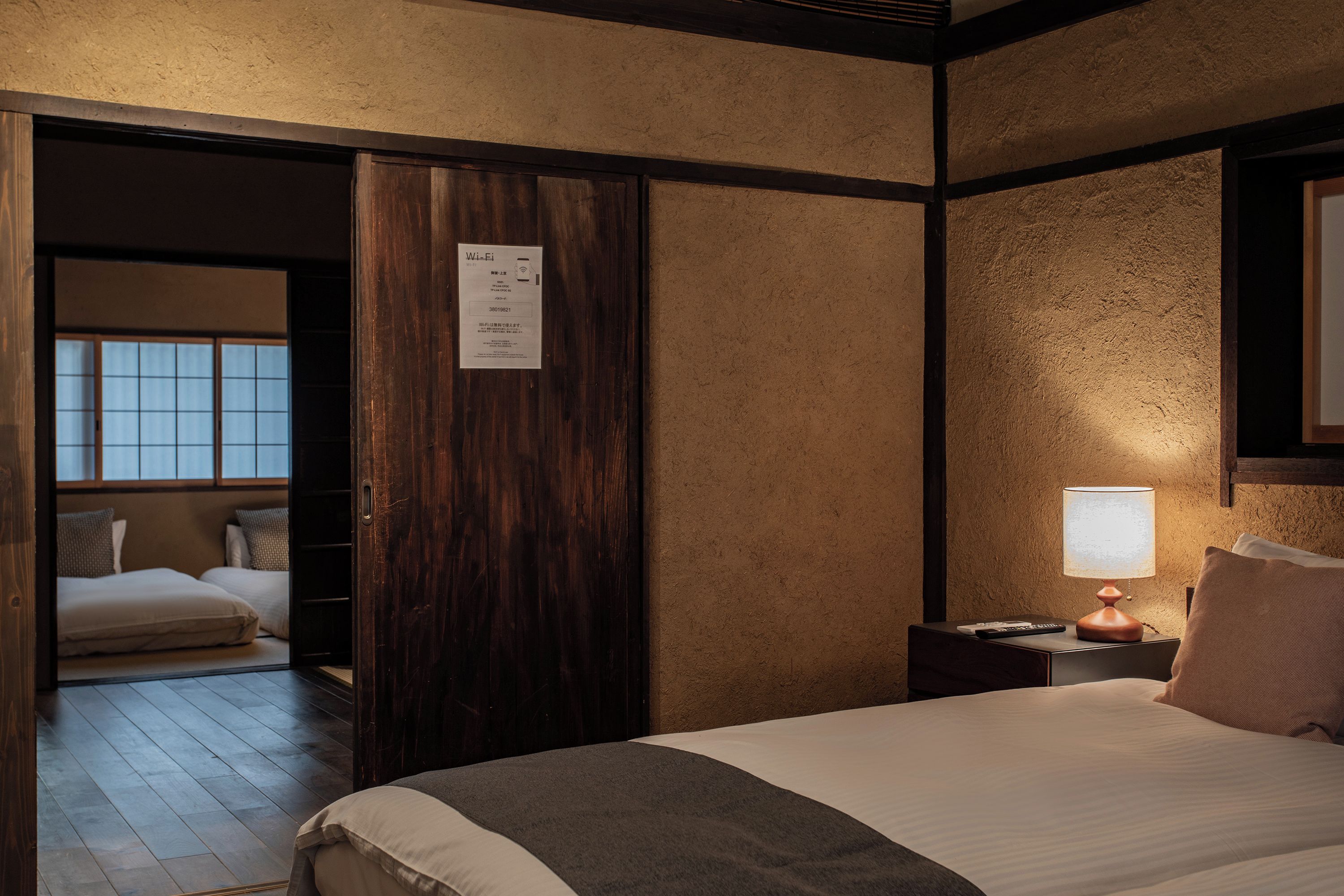 Quarto de hotel com cama, mesa de cabeceira, luminária e porta que dá para outro cômodo.