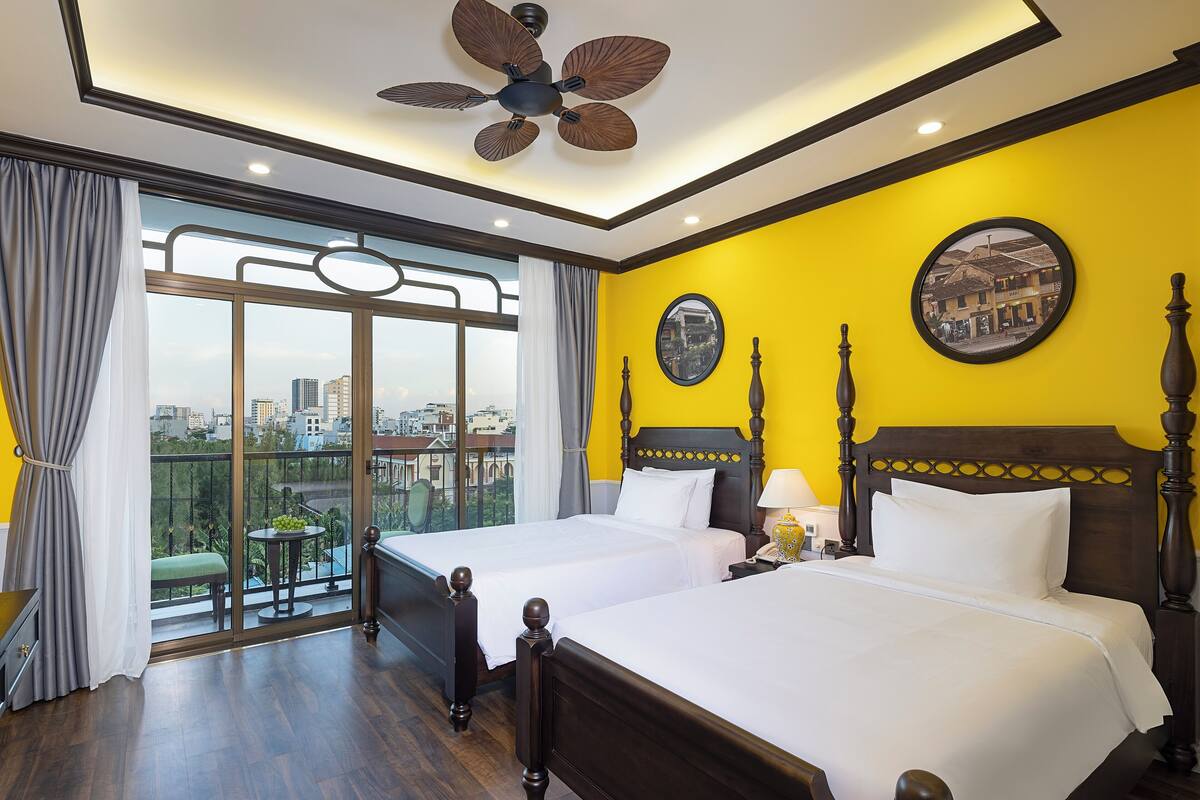 Quarto de hotel com duas camas, uma varanda e vista para a cidade, e um ventilador de teto.