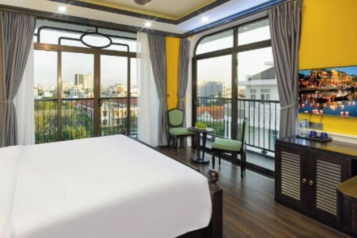 Quarto de hotel com uma cama grande, varanda com vista para a cidade, uma pequena mesa com cadeiras e uma televisão na parede.