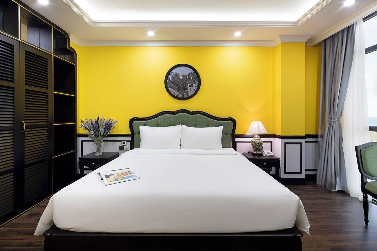 Quarto de hotel com uma cama grande, mesa de cabeceira, luminária, vaso com flores e um quadro na parede.
