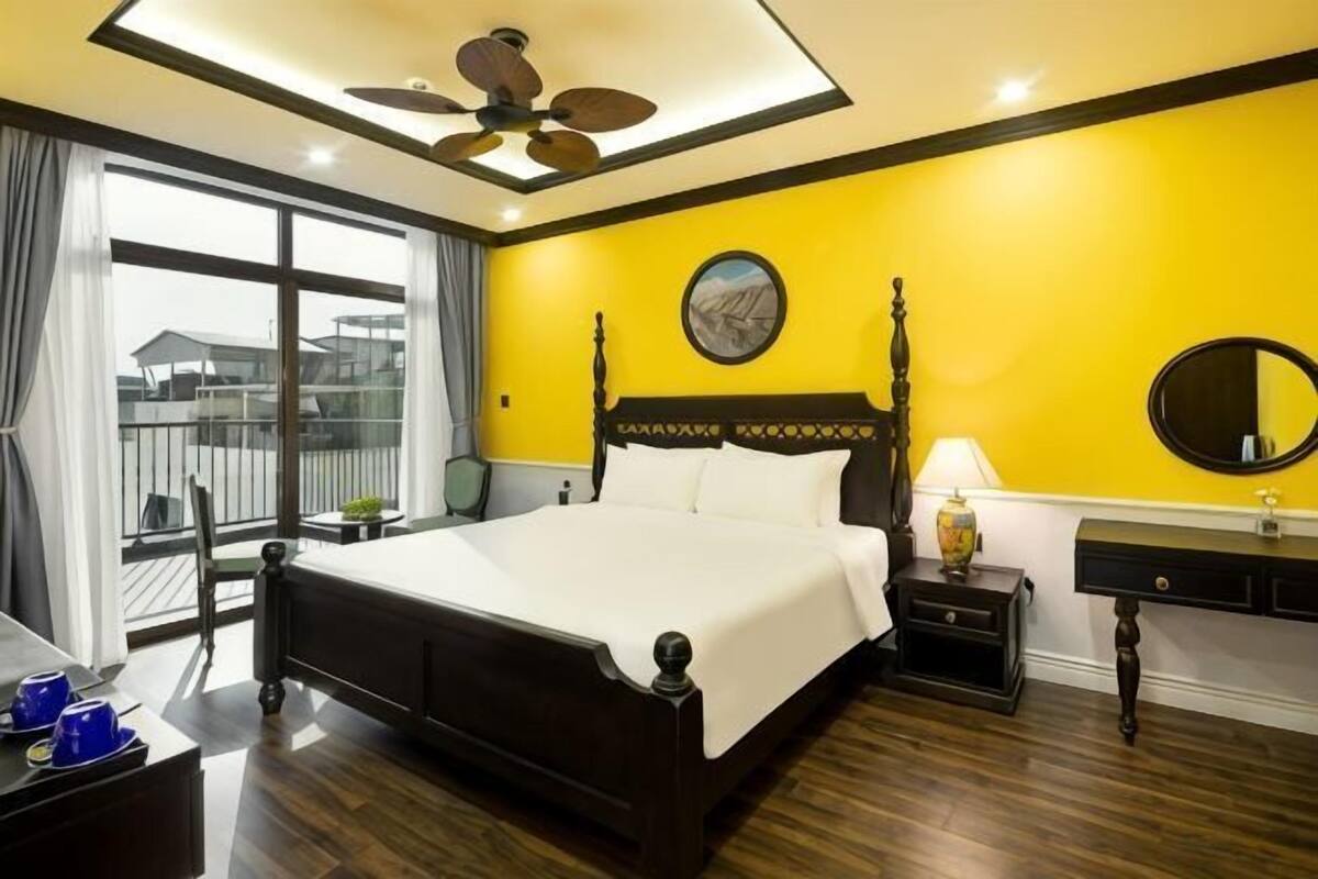 Quarto de hotel com uma cama grande, um ventilador de teto, uma janela com cortinas, uma mesa de cabeceira com abajur e um espelho na parede.