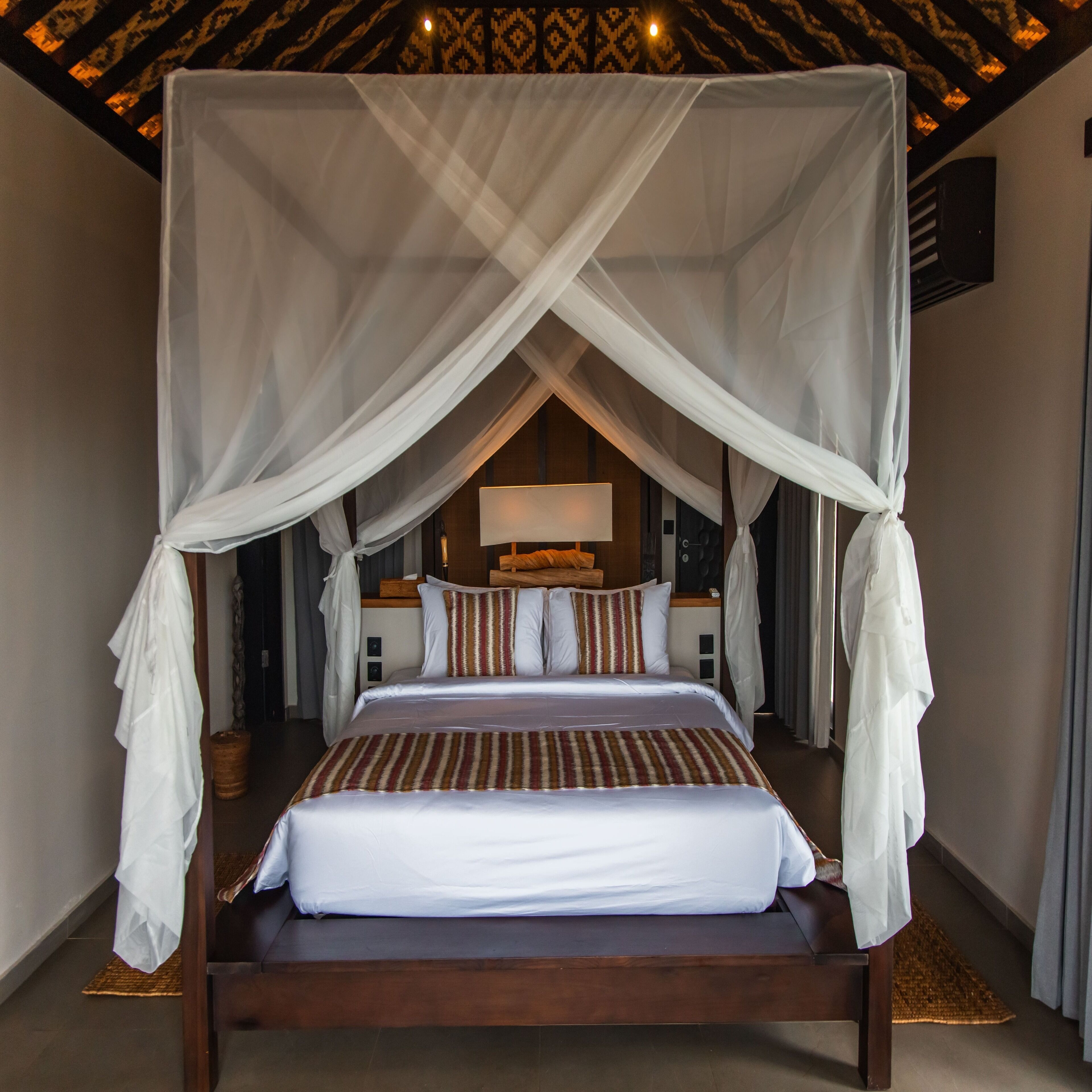 Uma cama com dossel e cortinas, roupa de cama branca, cabeceira de madeira e manta listrada.