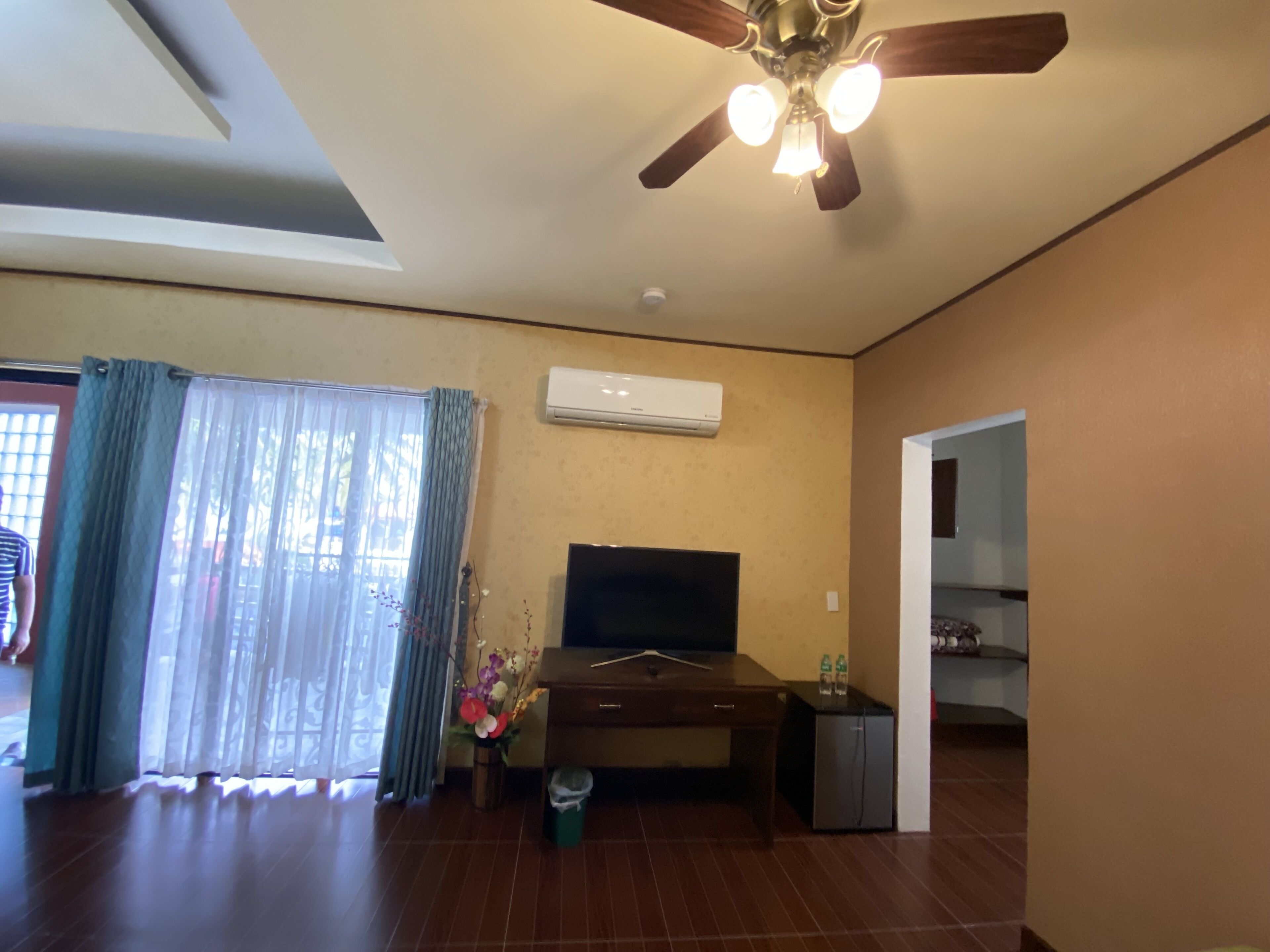 Uma sala de estar com ventilador de teto, televisão de tela plana, um frigorífico pequeno e uma janela com cortinas.