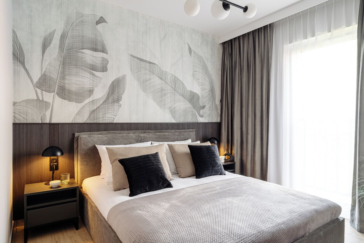 Um quarto moderno com uma cama grande, cabeceira com padrão de folha, duas mesas de cabeceira com luminárias e uma janela ampla com cortinas.