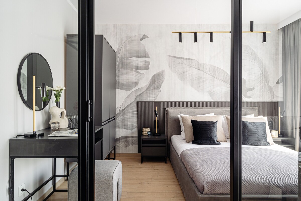 Um quarto moderno com uma cama grande, uma escrivaninha com um computador, um espelho e uma mesa de cabeceira com abajur.