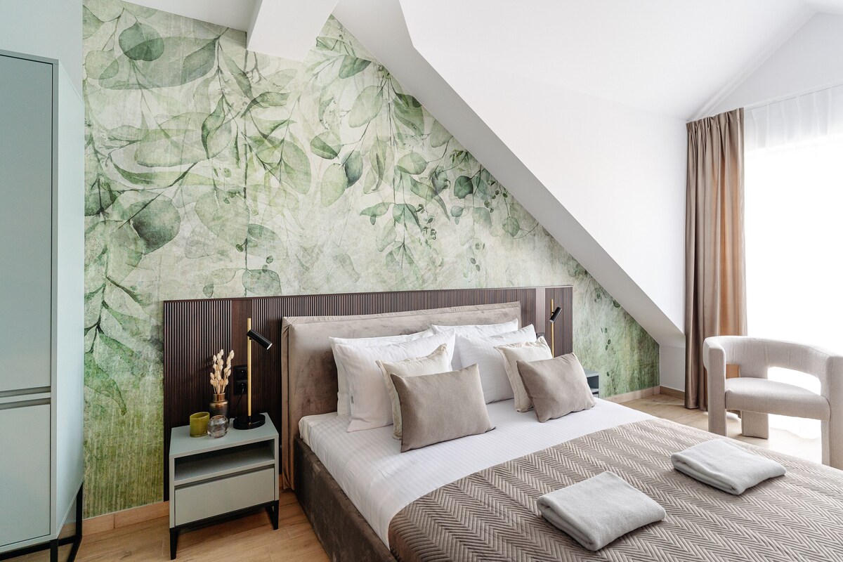 Quarto de hotel moderno com uma cama, uma mesa de cabeceira, uma cadeira e uma janela grande com cortinas.