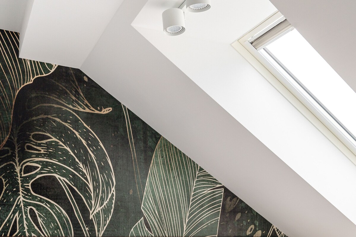 Quarto moderno com teto inclinado, uma cama grande com roupa de cama bege, um mural na parede e um claraboia.
