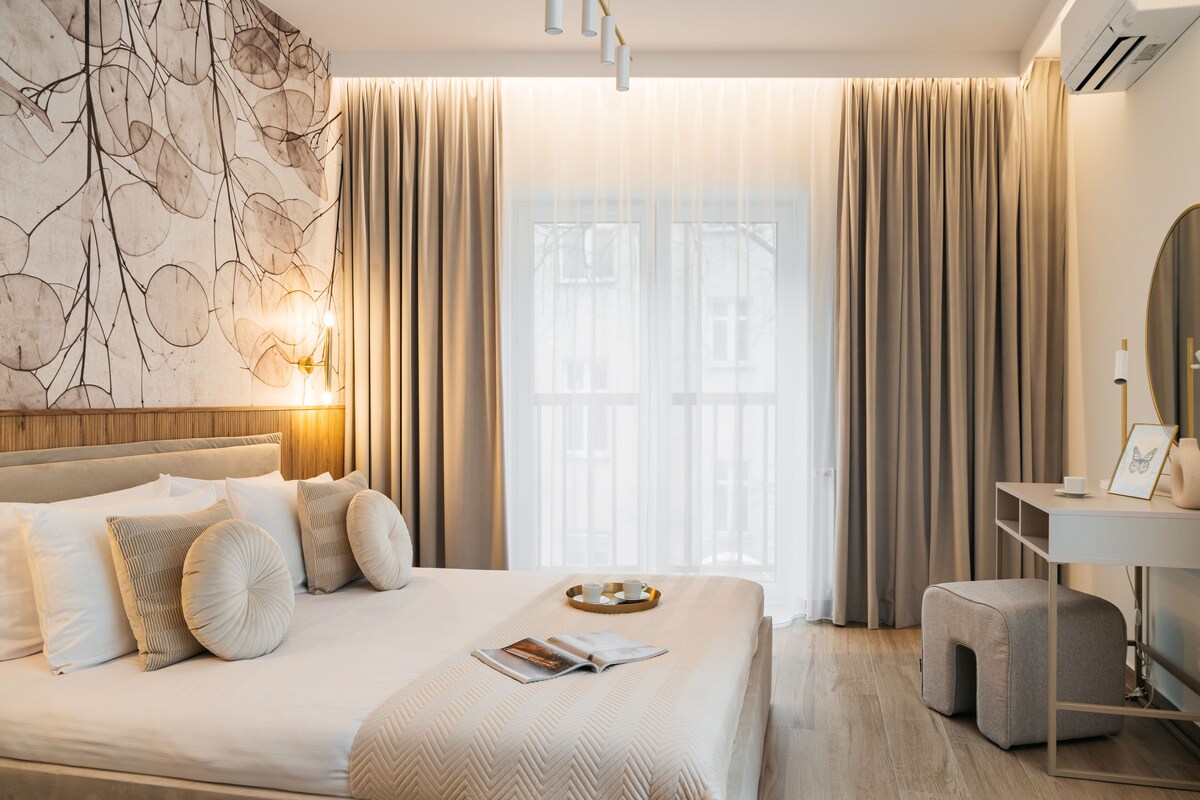 Quarto de hotel moderno com uma cama grande, mesa de cabeceira com abajur, um banquinho pequeno e vista da cidade por grandes janelas.