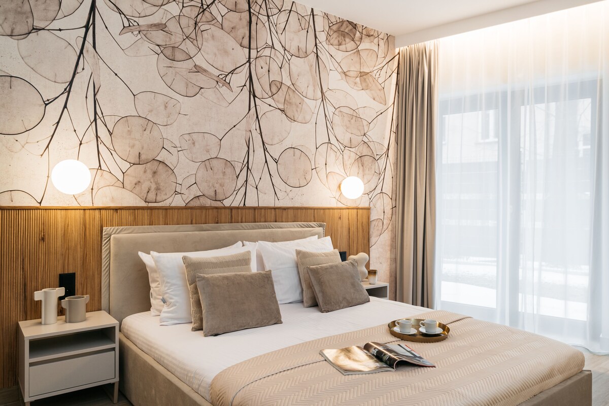 Quarto de hotel moderno com uma cama grande, uma mesinha de cabeceira, parede com padrão de folhagem e janela com cortinas.
