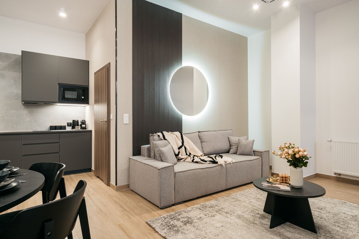 Uma sala de estar moderna com um sofá cinzento, uma mesa de centro redonda e uma área de cozinha com micro-ondas.