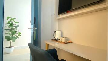 Quarto moderno com televisão de tela plana na parede, uma pequena mesa com chaleira e xícaras, uma cadeira azul e um vaso de plantas perto da porta.