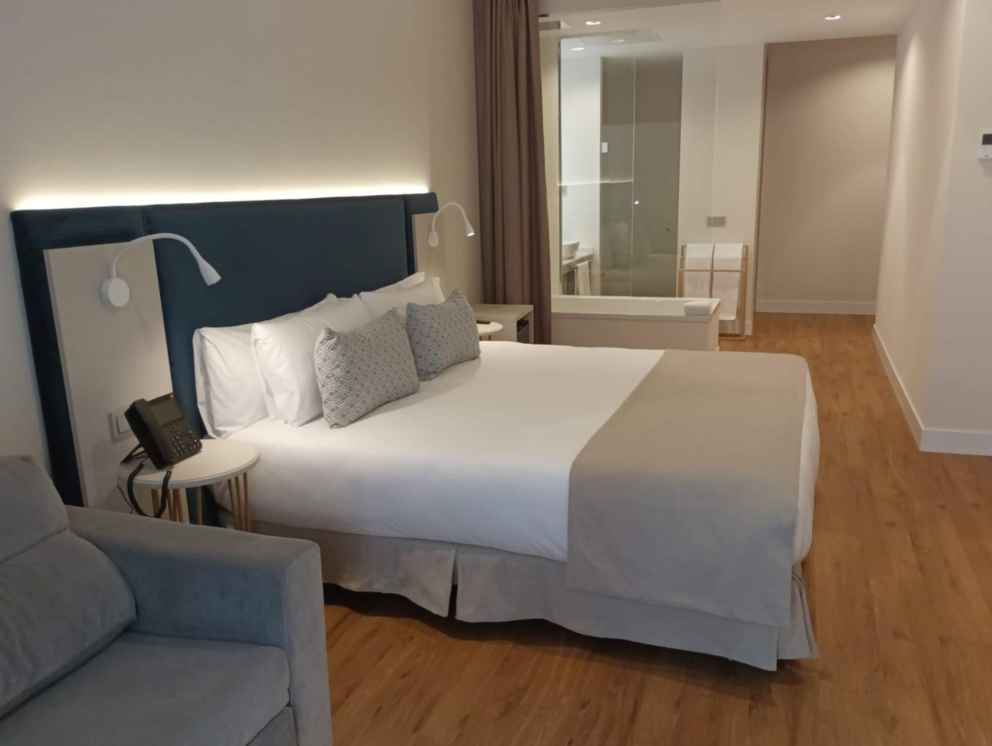 Quarto de hotel moderno com uma cama grande, mesas de cabeceira, telefone e um espaço de estar.