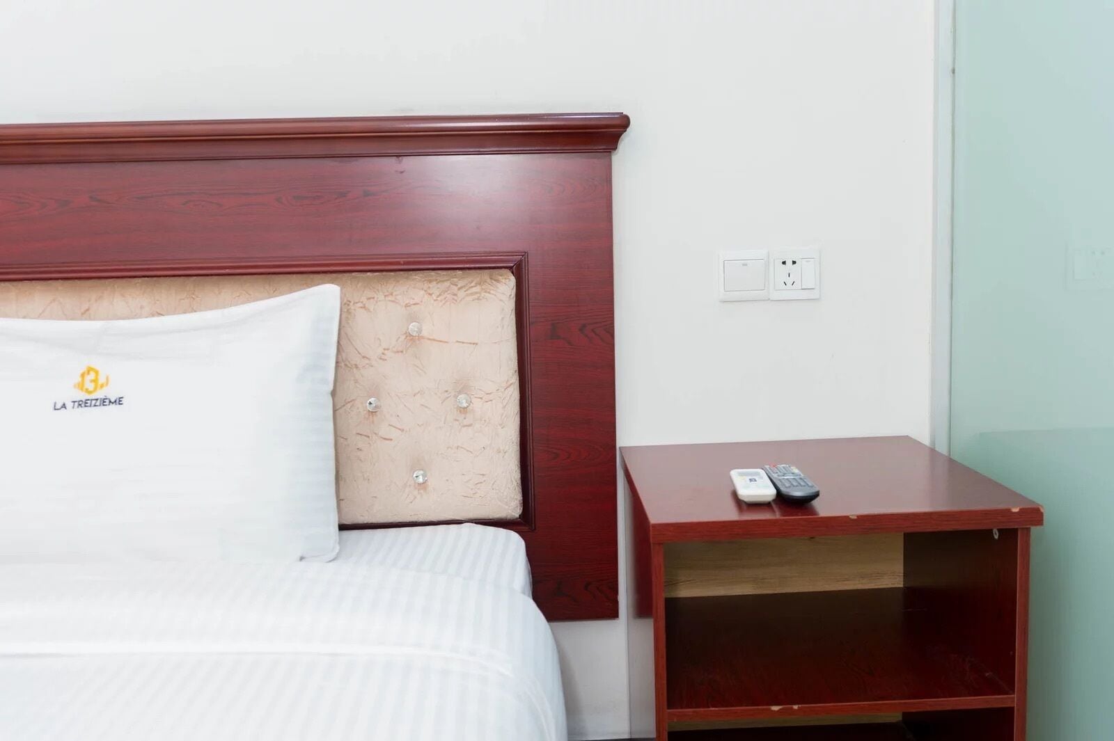 Quarto de hotel com cabeceira de madeira, travesseiro branco com logotipo, mesa de cabeceira com controle remoto e tomada na parede.