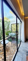 Quarto moderno com grandes portas de vidro que dão para um pátio com mesa e cadeira, rodeado por pedras e plantas.