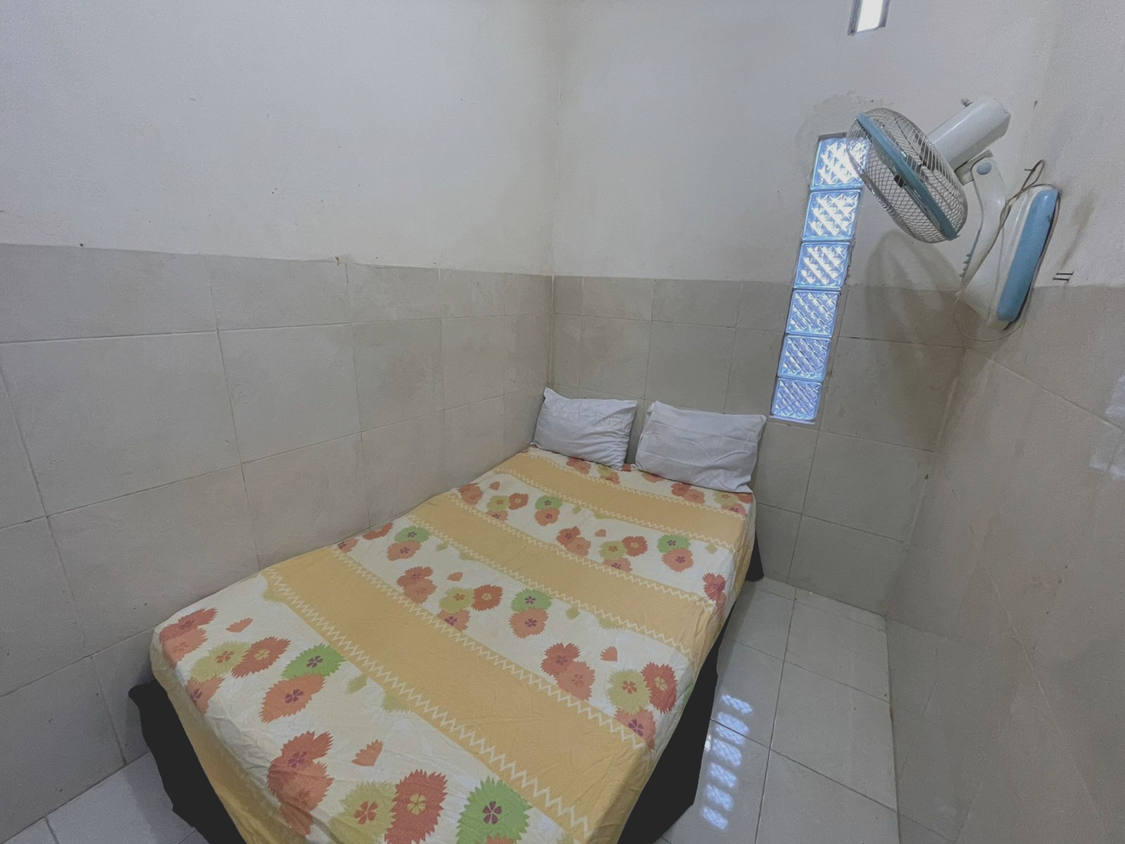 Quarto pequeno com uma cama de solteiro, colcha estampada com flores, um ventilador e uma janela com padrão quadriculado.