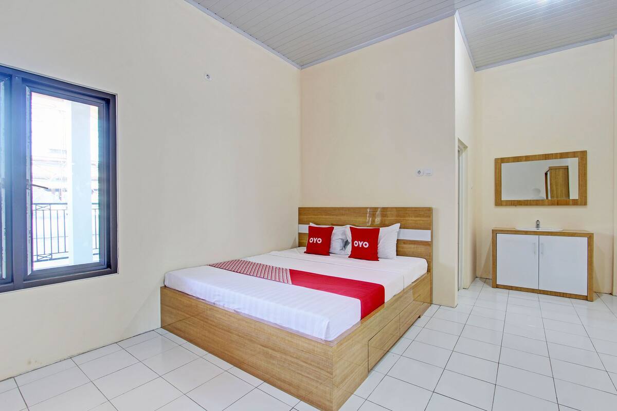 Um quarto com uma cama de madeira, roupa de cama branca, um conjunto de almofadas vermelho e branco, um pequeno armário de madeira e uma janela com vista para edifícios.