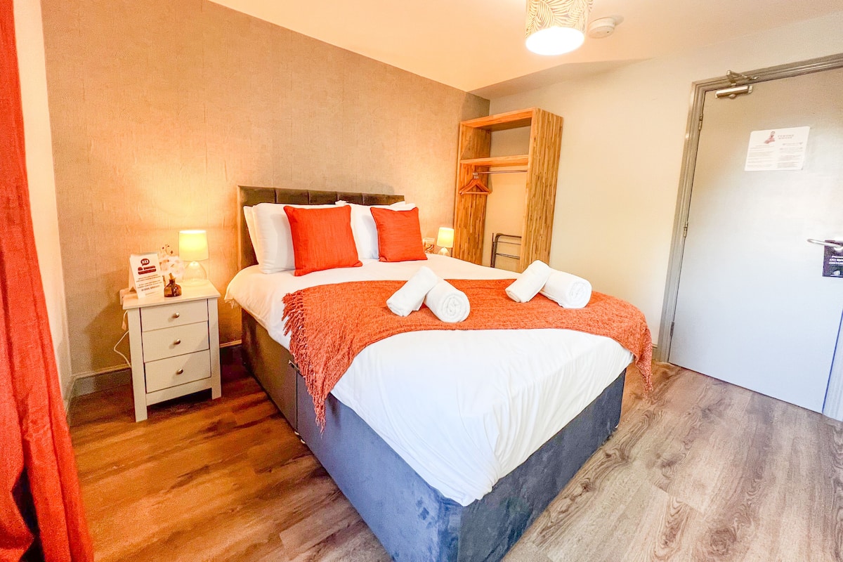Cama bem arrumada com roupa de cama branca e laranja, cabeceira de madeira e mesa de cabeceira com abajur.