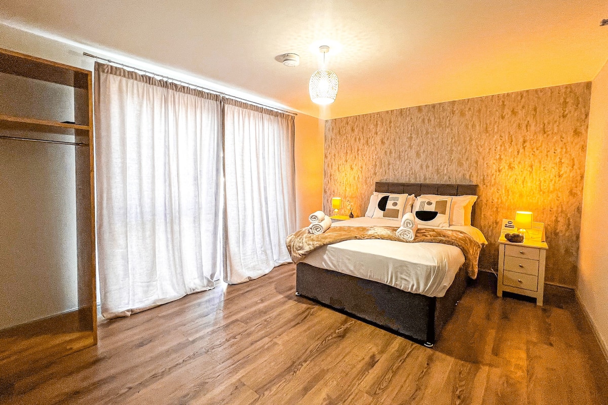 Quarto de hotel com piso de madeira, cama com cabeceira, mesa de cabeceira com abajur e guarda-roupa.