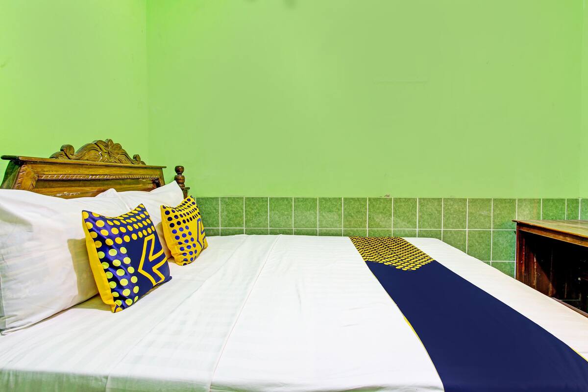 Cama bem arrumada com roupa de cama branca e travesseiros com padrão azul e amarelo, diante de uma parede com azulejos verdes.