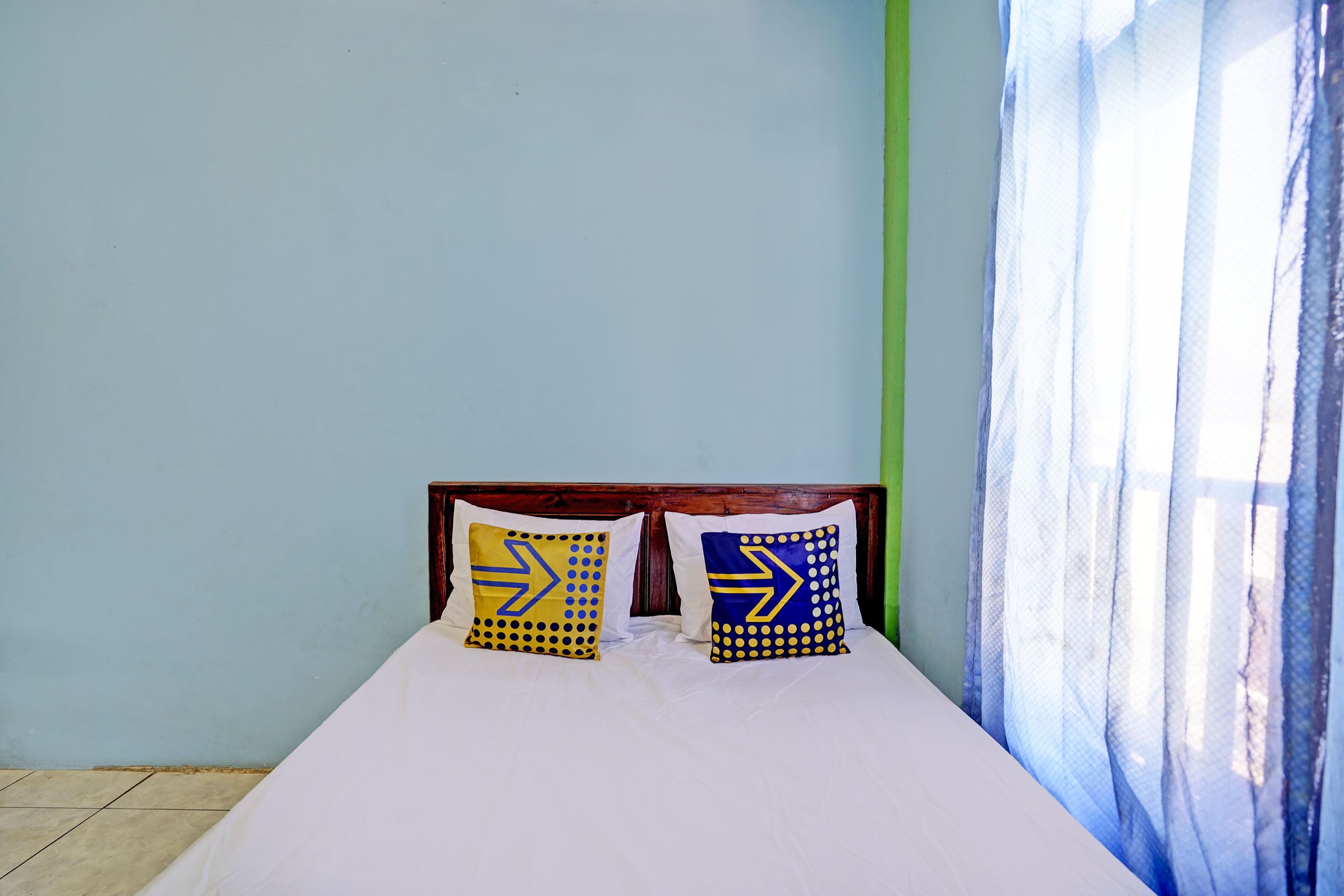 Uma cama com duas almofadas, cabeceira de madeira e janela com cortinas finas.