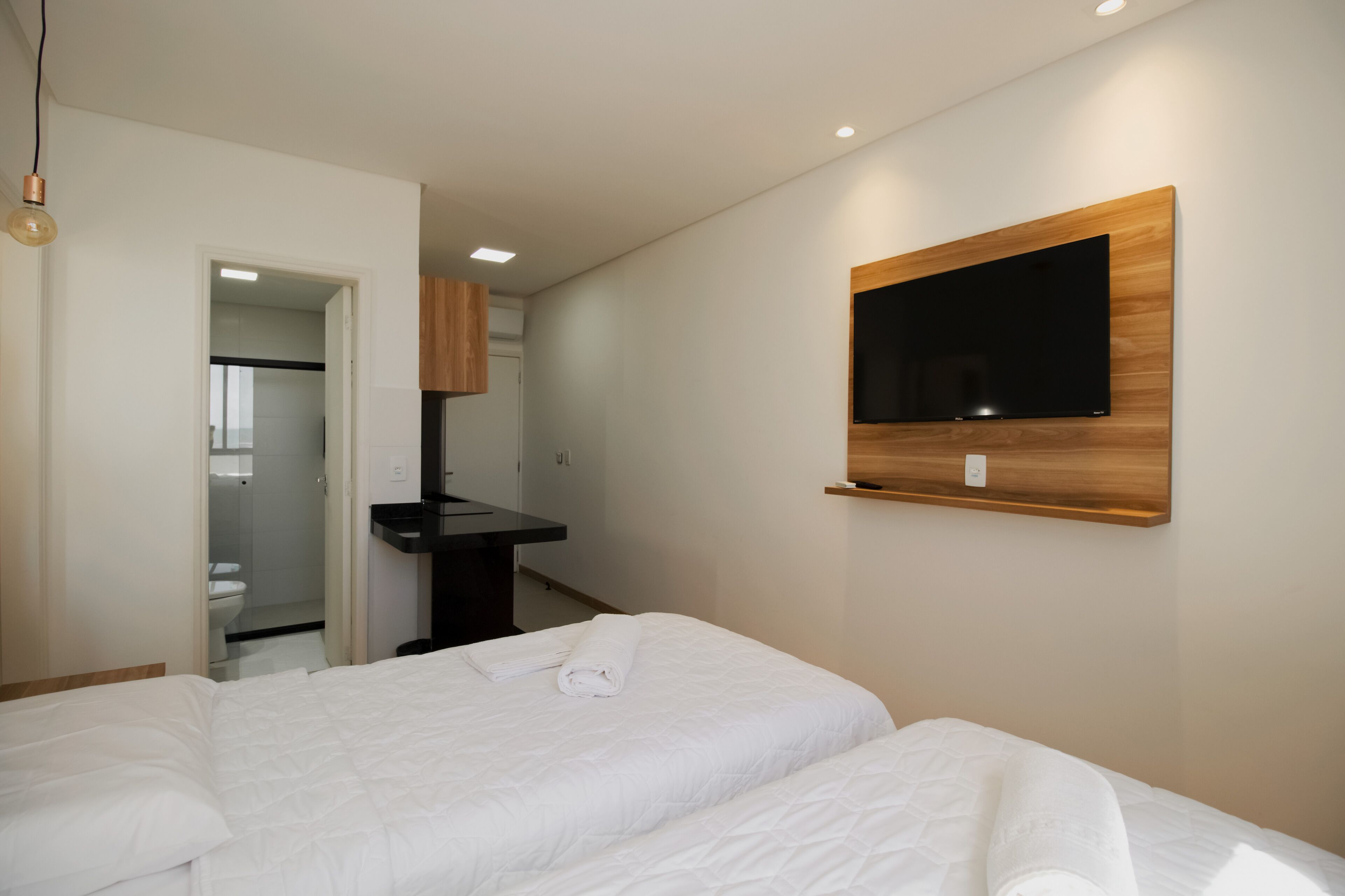 Quarto de hotel com duas camas, uma estante de madeira para a televisão, banheiro com vaso sanitário e chuveiro, e uma pequena escrivaninha com espelho.