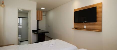 Quarto de hotel com duas camas, uma estante de madeira para a televisão, banheiro com vaso sanitário e chuveiro, e uma pequena escrivaninha com espelho.