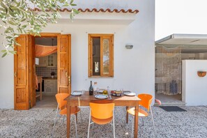 Uma área de refeições ao ar livre com mesa de madeira e cadeiras laranjas, cozinha visível por uma porta aberta e uma parede externa branca.