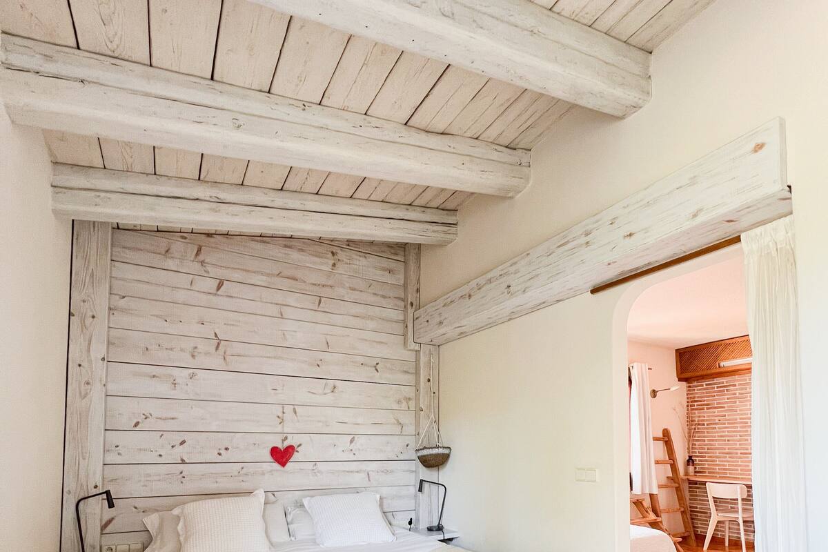 Quarto com teto de madeira, cama com roupa de cama branca, mesa de cabeceira com abajur e parede de tijolos visível por uma porta aberta.