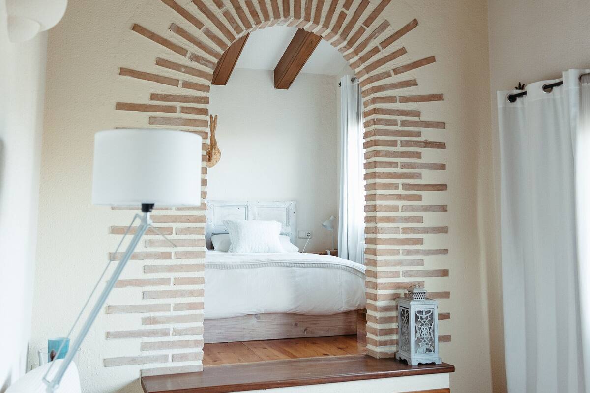 Uma escadaria leva a um quarto com uma cama branca, uma luminária e um pequeno candeeiro.
