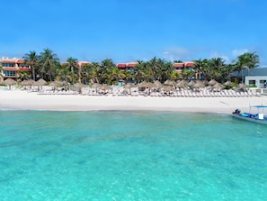 Resort à beira-mar com águas turquesa cristalinas, areia branca e fofa, fileiras de espreguiçadeiras, palmeiras e um barco.